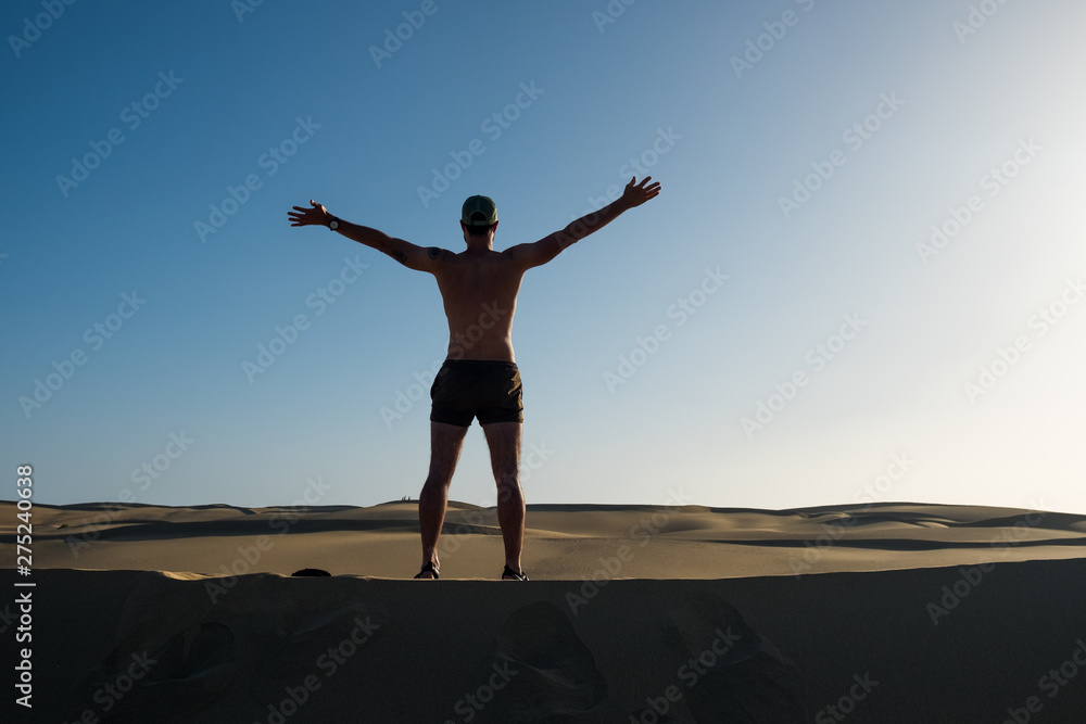 young man standing on top of sand desert dune with open arms as a ...