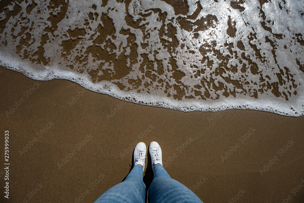 Woman feet view with jeans from above at the beach with sand and wave ...