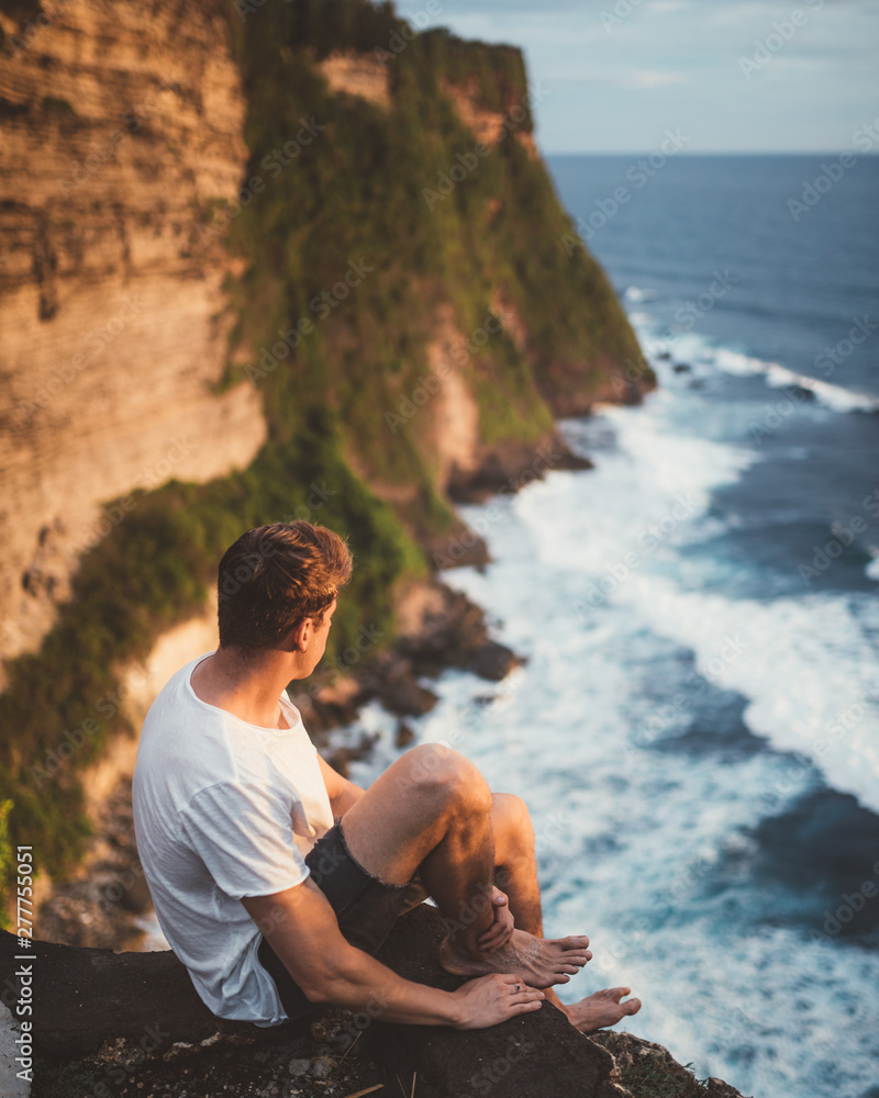 Side view of man sitting alone on cliff at height looking at majestic ...