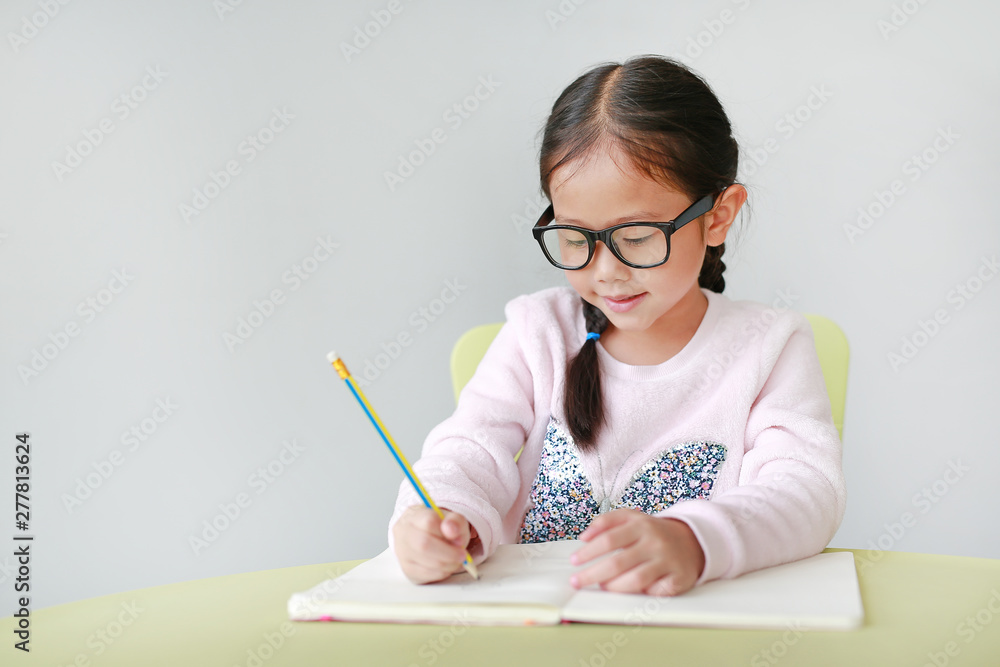 Smiling little Asian child girl wearing eyeglasses and write in a book ...