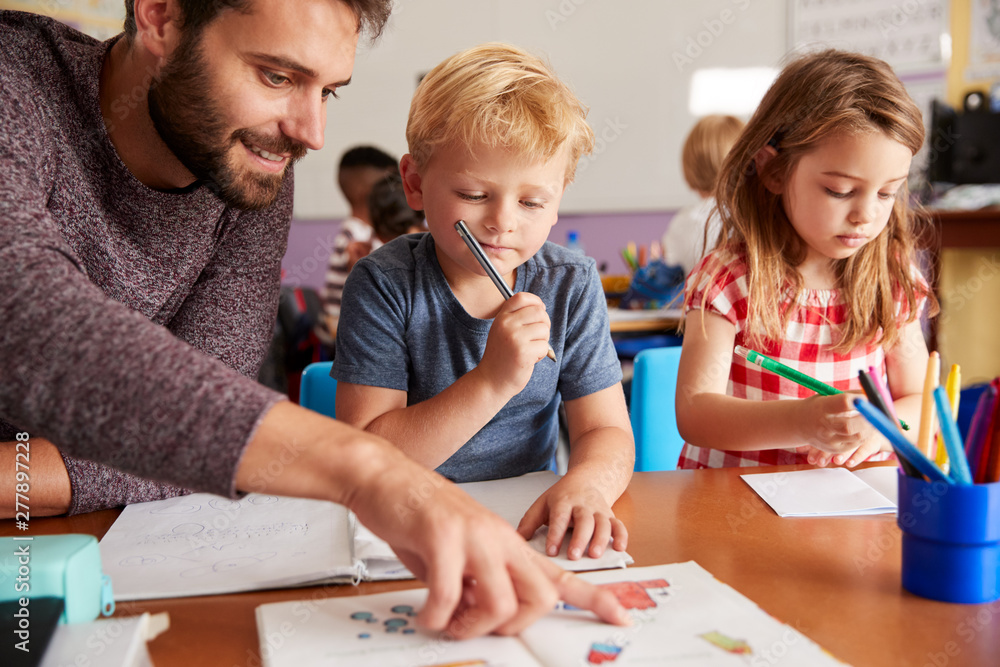 Elementary School Teacher Helping Pupils As They Work At Desk In ...