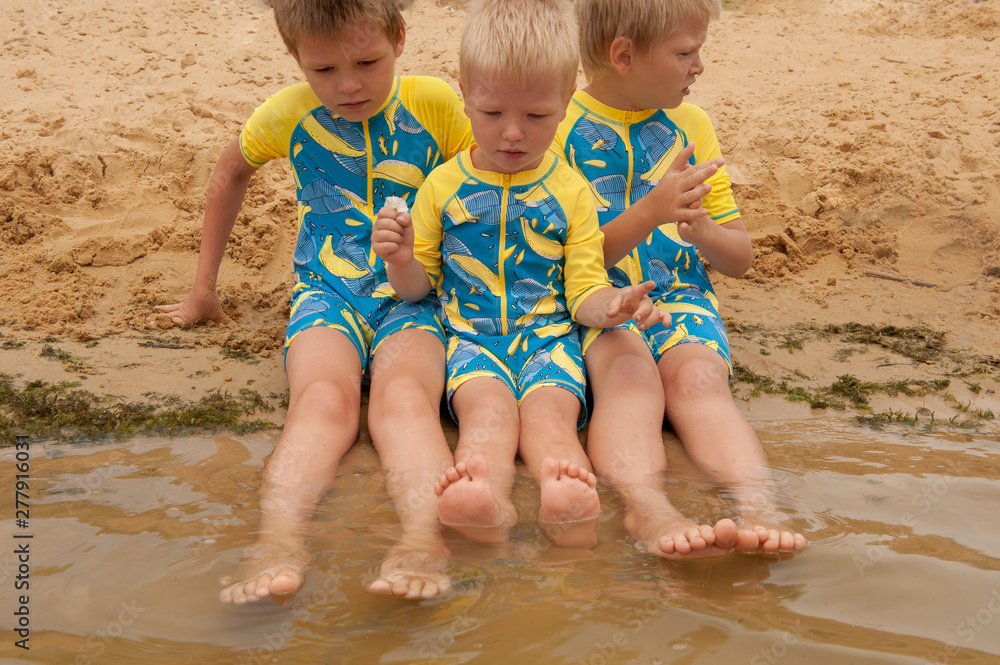 Funny kids in bathing suits sit together on sand with their legs in ...