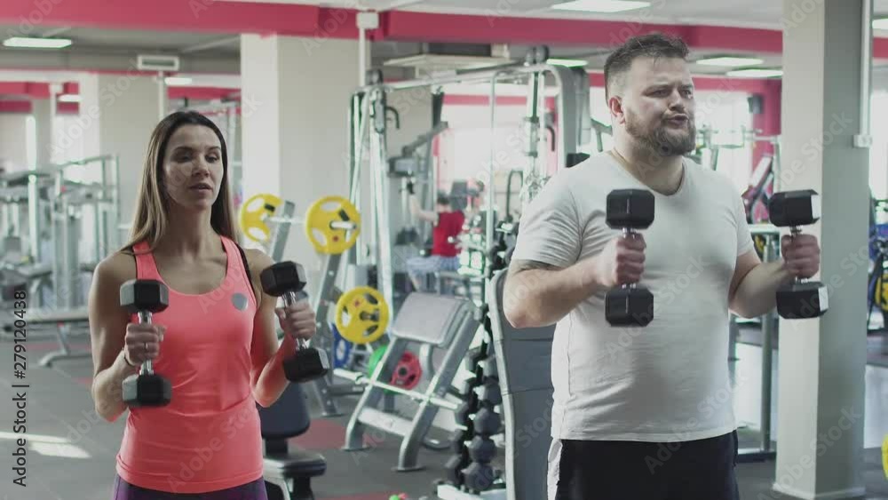 Thick obese guy with individual coach do physical jerks with dumbbells ...