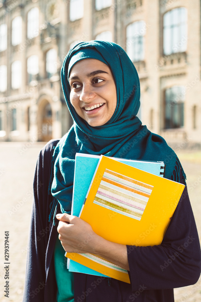 Smiling muslim young female student holding books standing near college ...