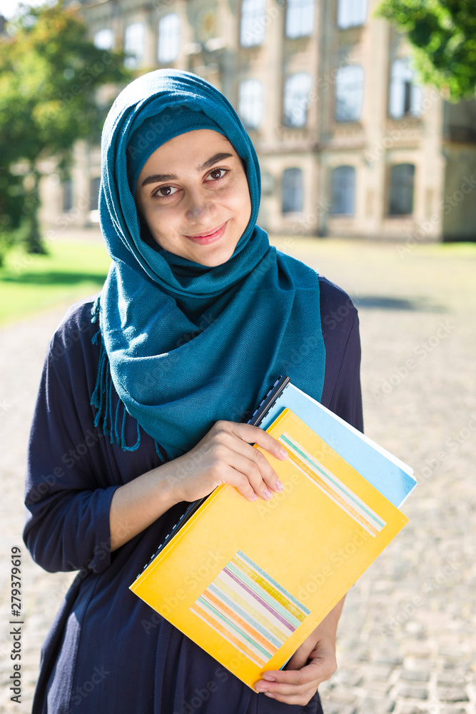 Smiling muslim young female student holding books standing near college ...