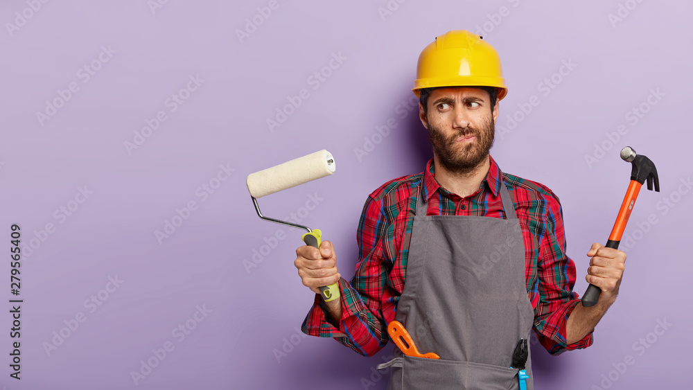 Discontent male builder frowns face, wears protective helmet, holds ...