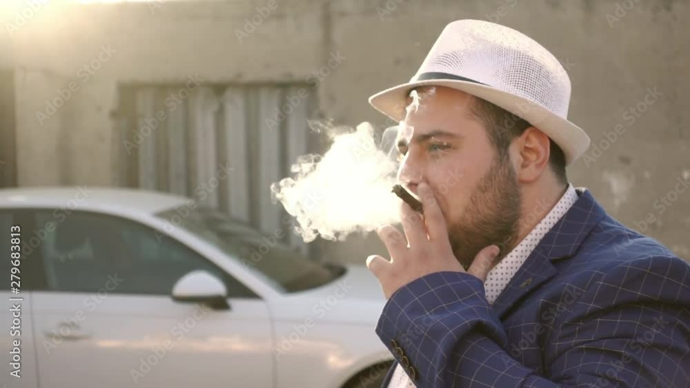 Portrait of bearded young man in hat and suit smokes a cigar in parking ...