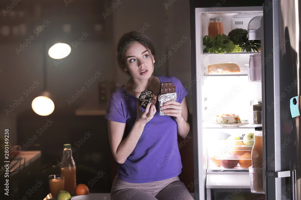Wary young woman eating unhealthy food near fridge in kitchen at night ... Wary young woman eating unhealthy food near fridge in kitchen at night ...
