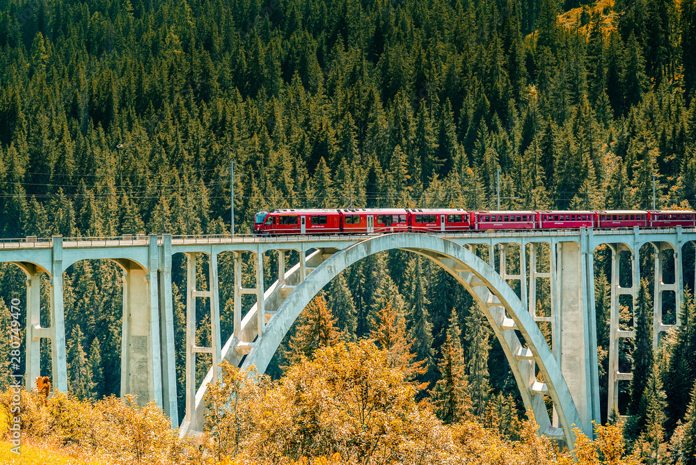 red train crosses a long viaduct across a deep ravine in the Swiss Alps ...
