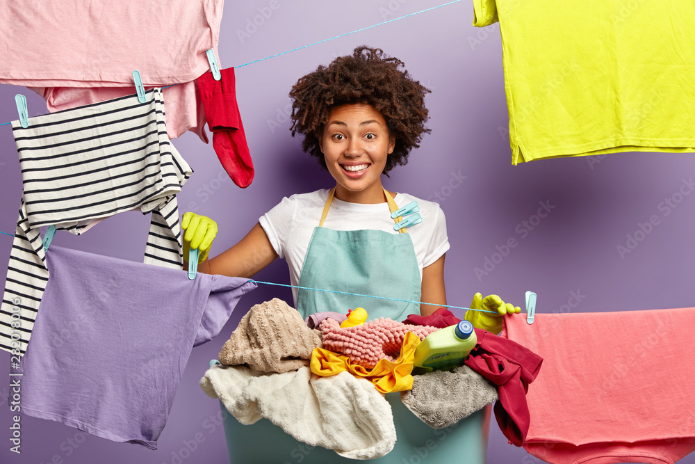 Smiling dark skinned housekeeper hangs clean clothes with clothespegs ...