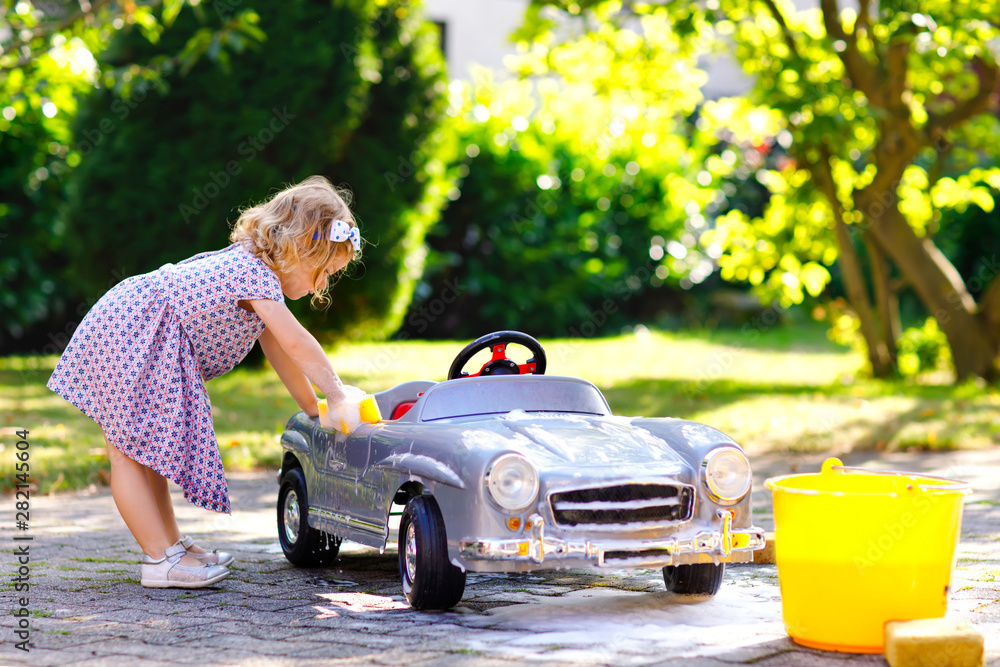 Cute gorgeous toddler girl washing big old toy car in summer garden ...