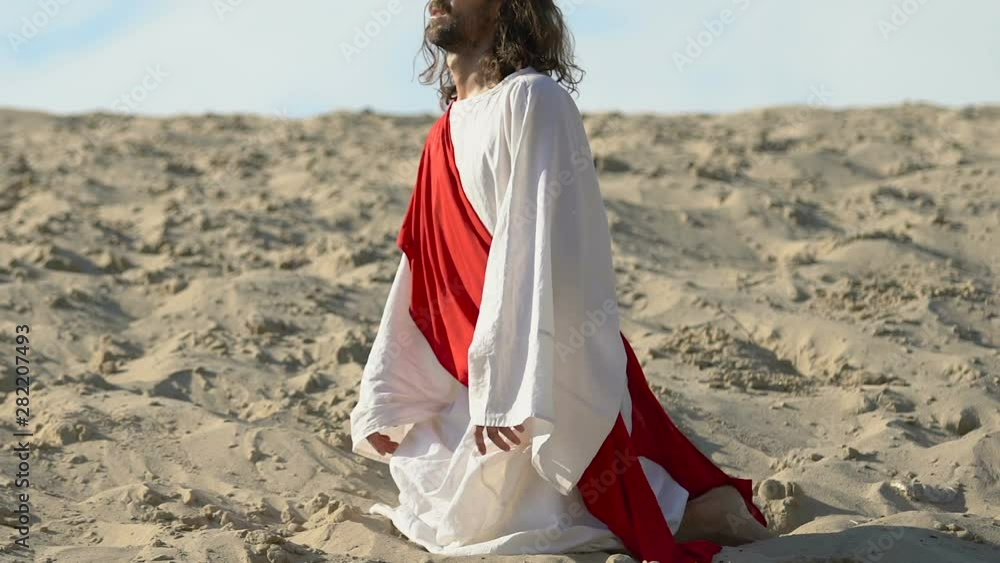 Exhausted Jesus in robe falling on knees in desert, raising hands to ...