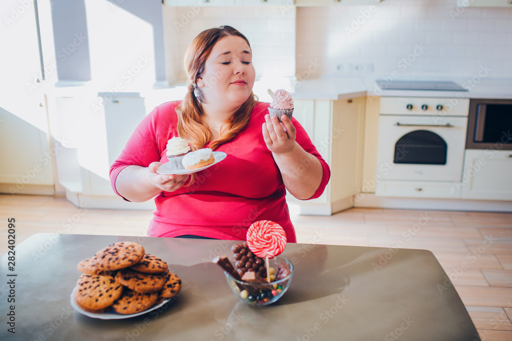 Fat young woman in kitchen sitting and eating sweet food. Plus size ...