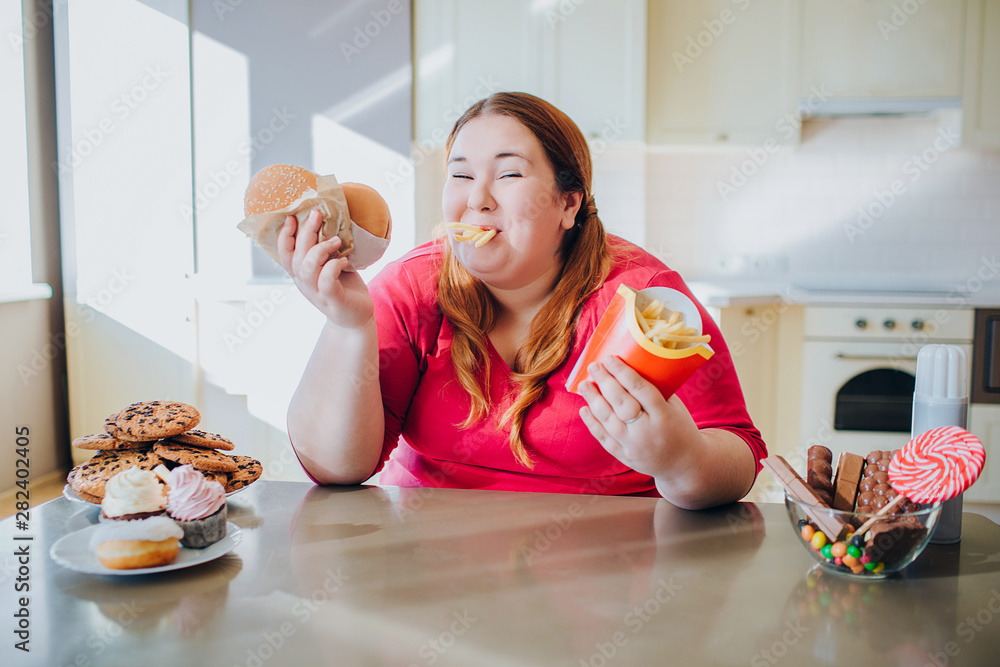 Fat young woman in kitchen sitting and eating junk food. French fries ...