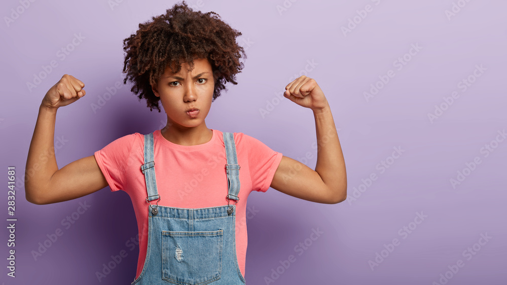 Serious looking strong woman shows muscles and biceps, scowls and looks ...