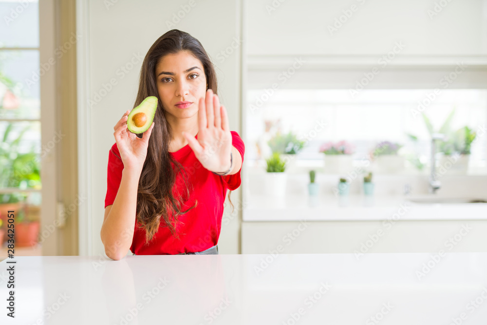 Young woman eating healthy avocado with open hand doing stop sign with ...
