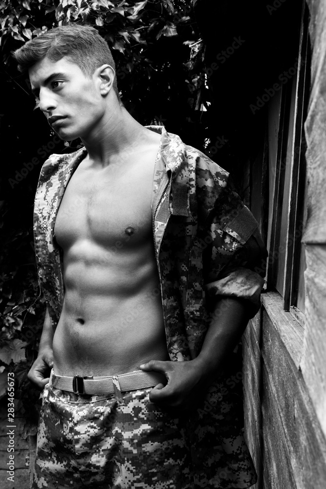 Good looking male soldier in uniform standing next wooden hut shows off ...