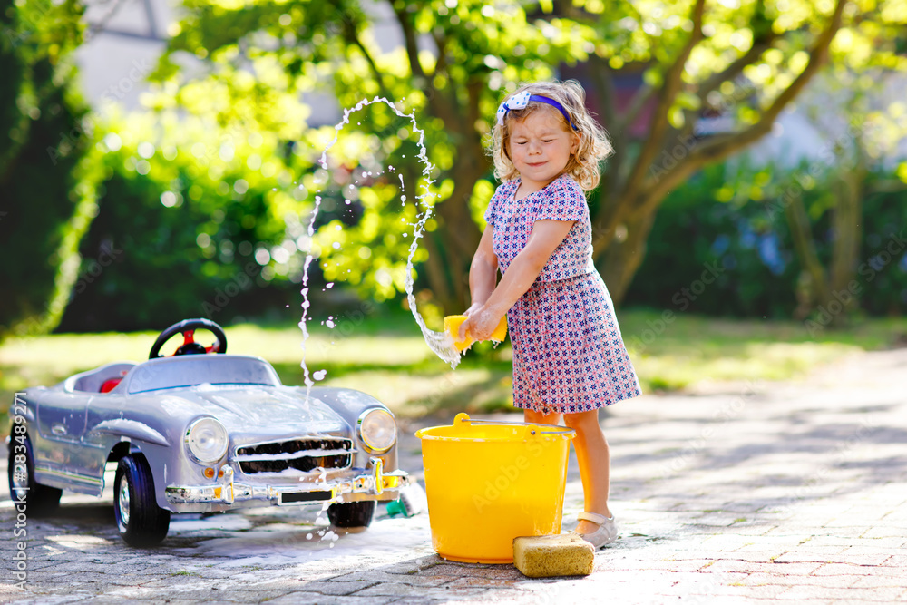 Cute gorgeous toddler girl washing big old toy car in summer garden ...