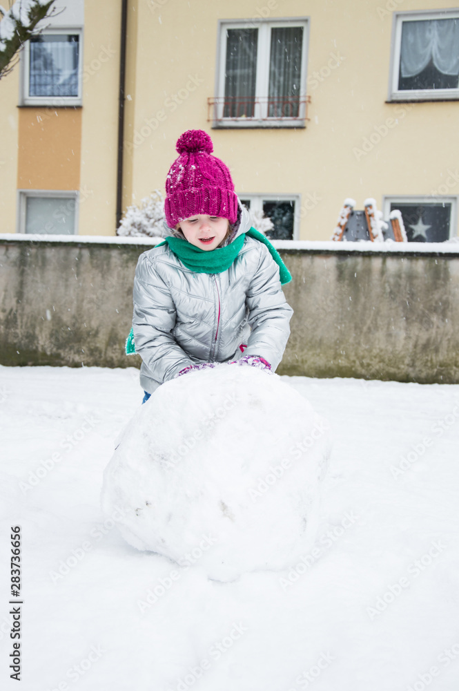 Cute little girl is rolling snowball. children make snowman in the yard ...