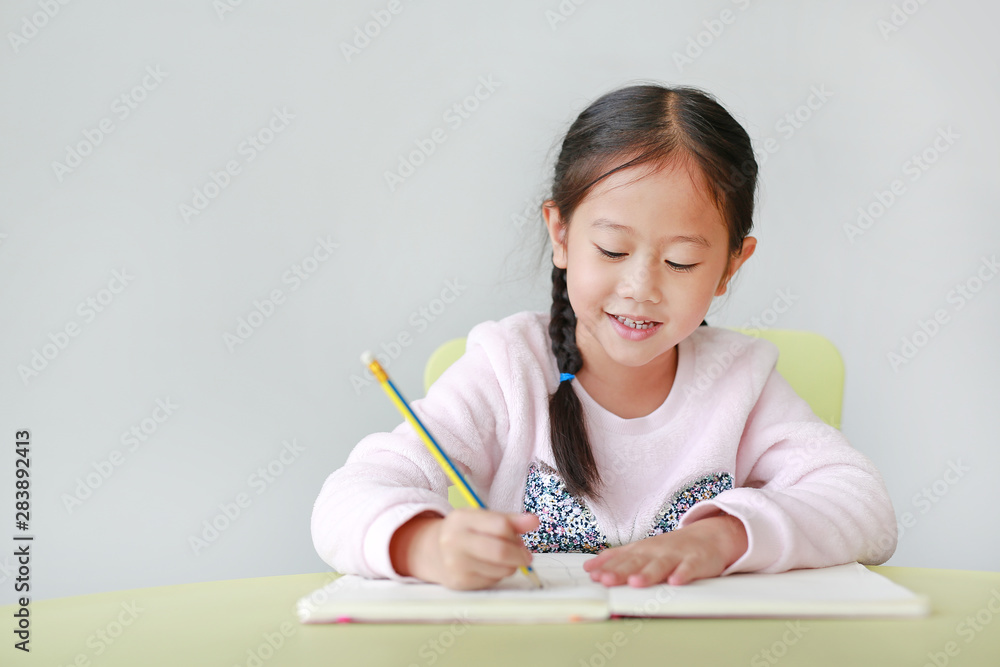 Happy little Asian child girl writes in a book or notebook with pencil ...