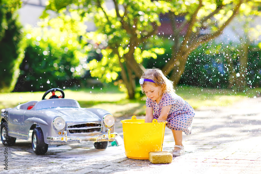 Cute gorgeous toddler girl washing big old toy car in summer garden ...