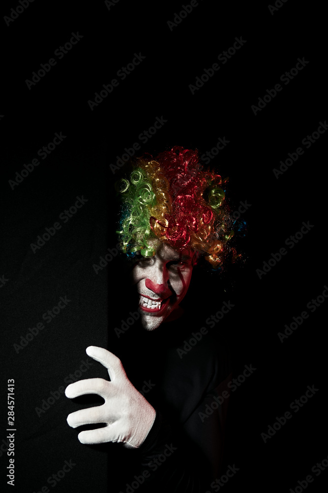 Scary clown peeping around the corner of a black wall. He is wearing a ...