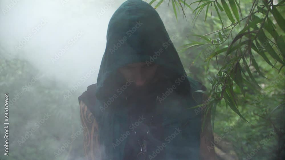 Man wearing hood standing in a dark tropical jungle and smoke billowing ...