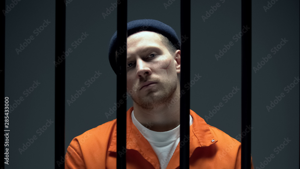 Dangerous criminal with scared face looking at camera behind prison ...