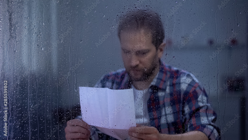 Sad man reading letter behind rainy window, bank credit payment overdue ...