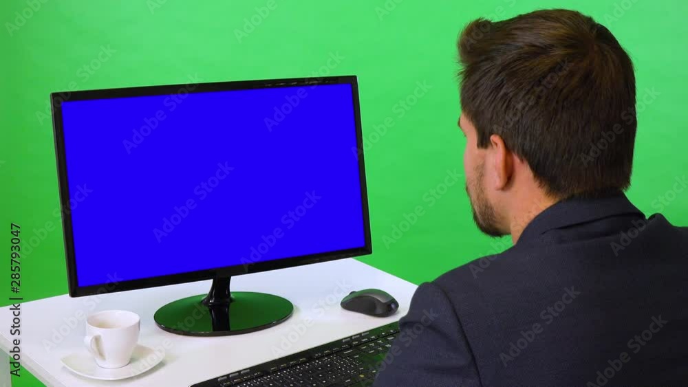 A young businessman sits at a desk and looks at a desktop computer with ...