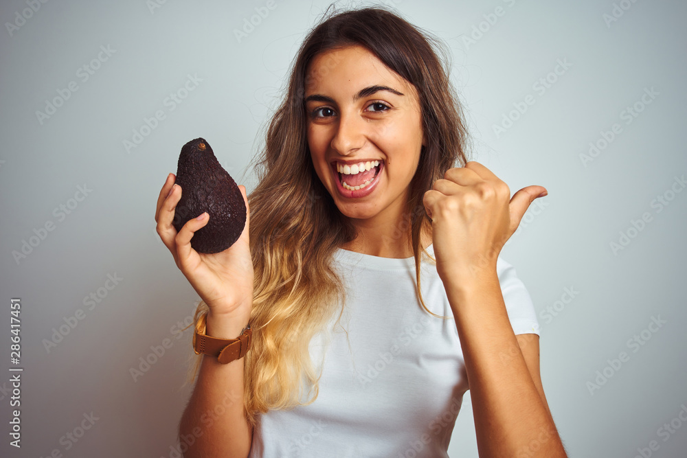 Young beautiful woman eating avocado over grey isolated background ...