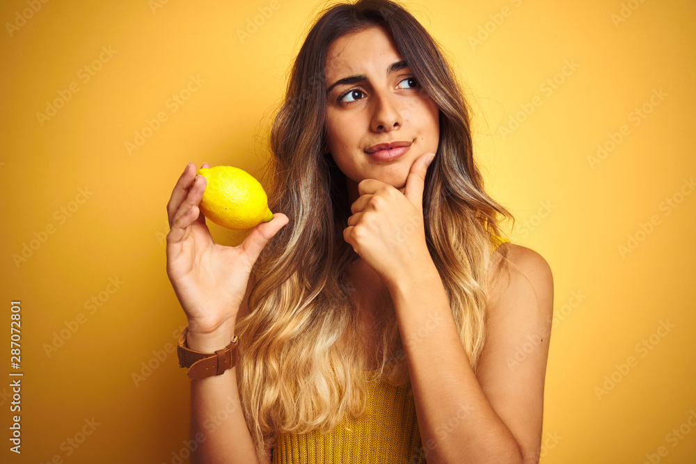 Young beautiful woman eating a lemon over yellow isolated background serious face thinking about ...