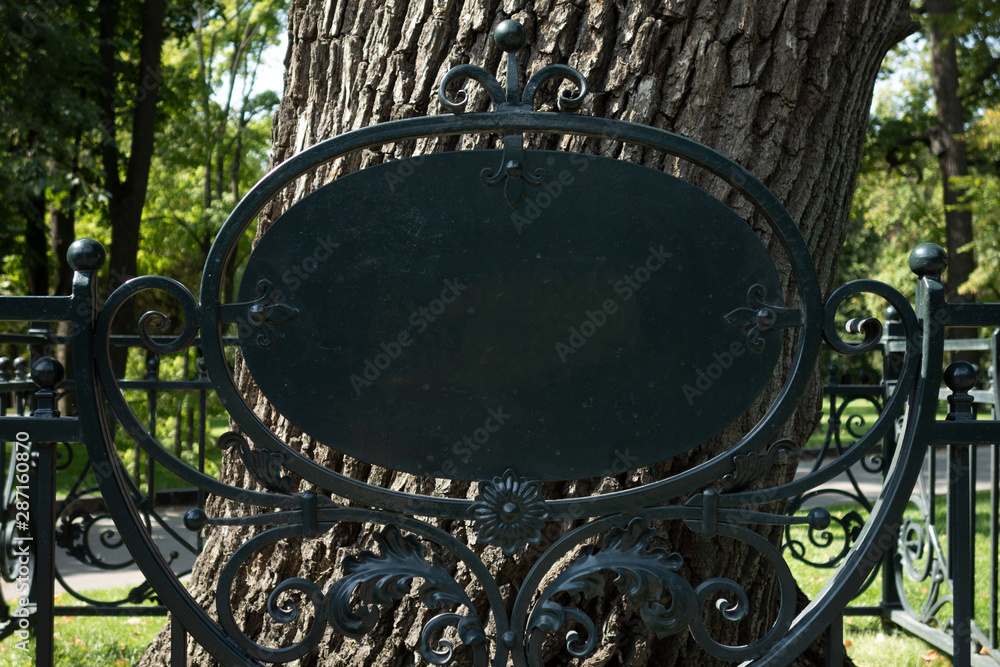 Empty blank sign on metal forging fence in front of big tree. Mock up ...