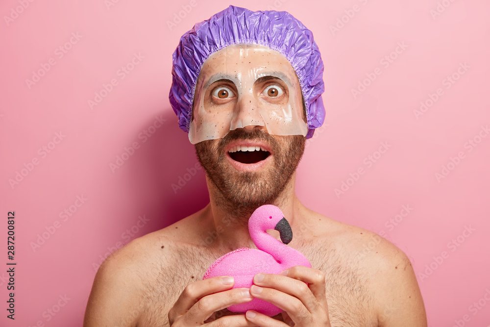 Surprised young man applies cosmetic mask holds bath sponge in shape of ...