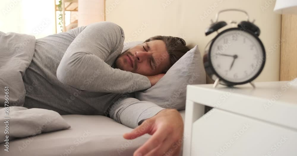 Sleepy man turning alarm clock off and going back to sleep in the ...
