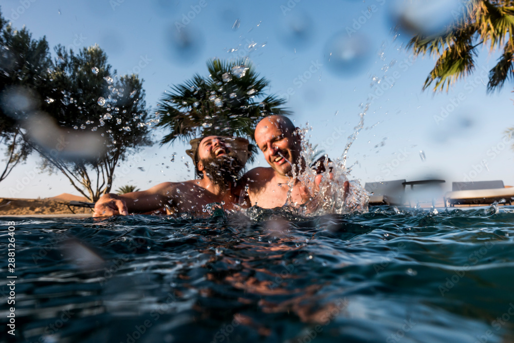 Gay couple in the pool next to palm trees and desert dunes, splashing ...