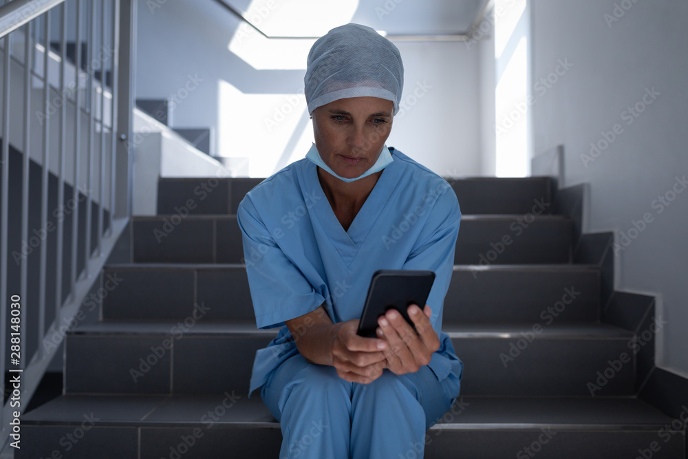 Female surgeon using mobile phone while sitting on hospital stair case ...