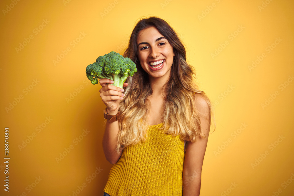 Young beautiful woman eating broccoli over yellow isolated background ...