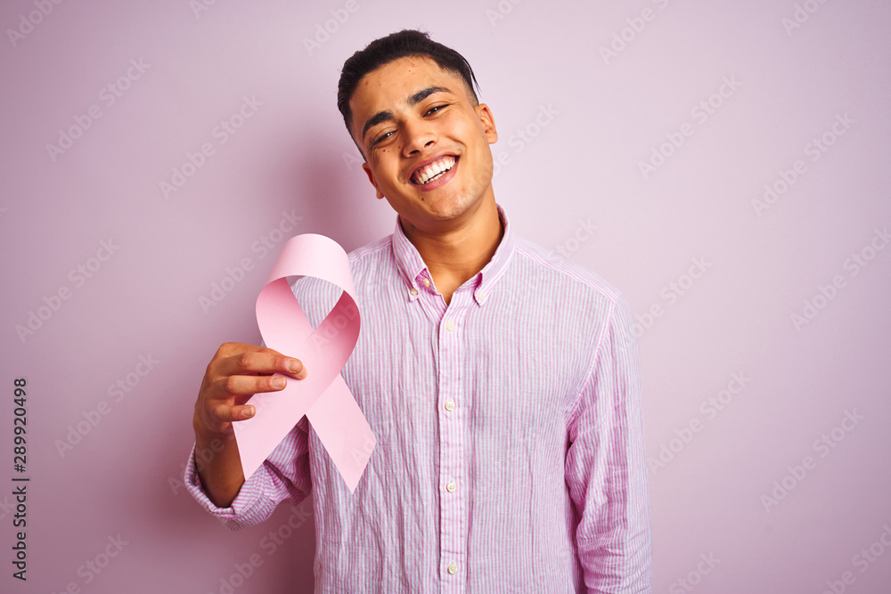 Young brazilian man holding cancer ribbon standing over isolated pink ...