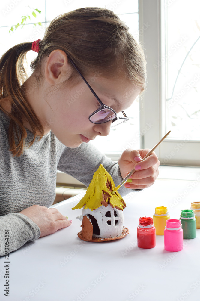 Girl making toys with your own hands, paints a clay house with gouache ...