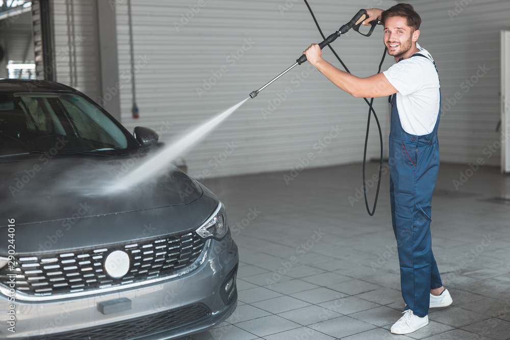 young handsome man wearing uniform washing car at car washing station ...
