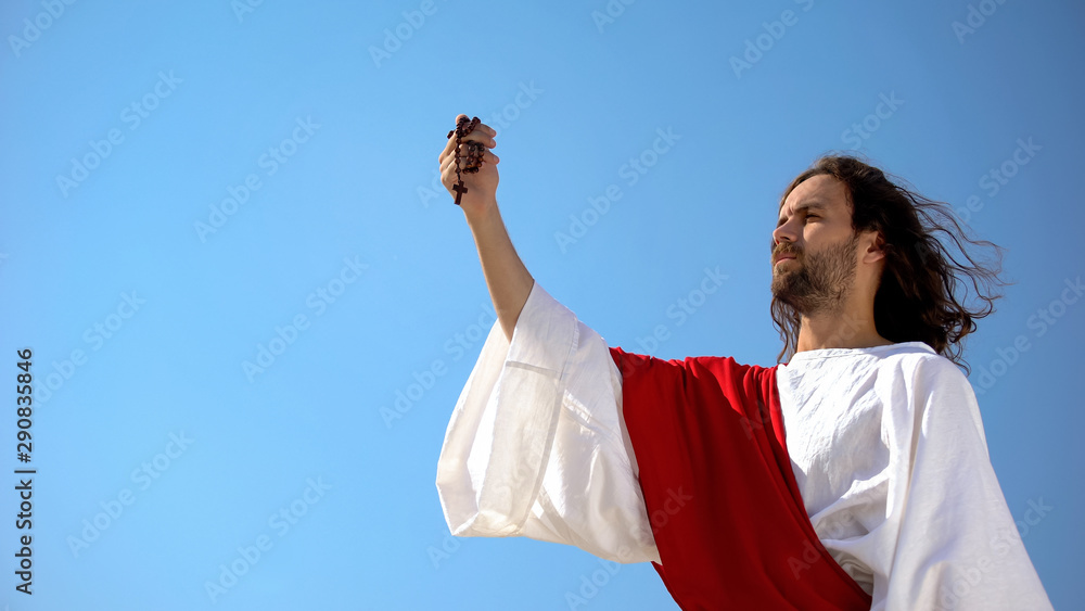 Righteous man raising hand with rosary against blue sky, praying to God ...