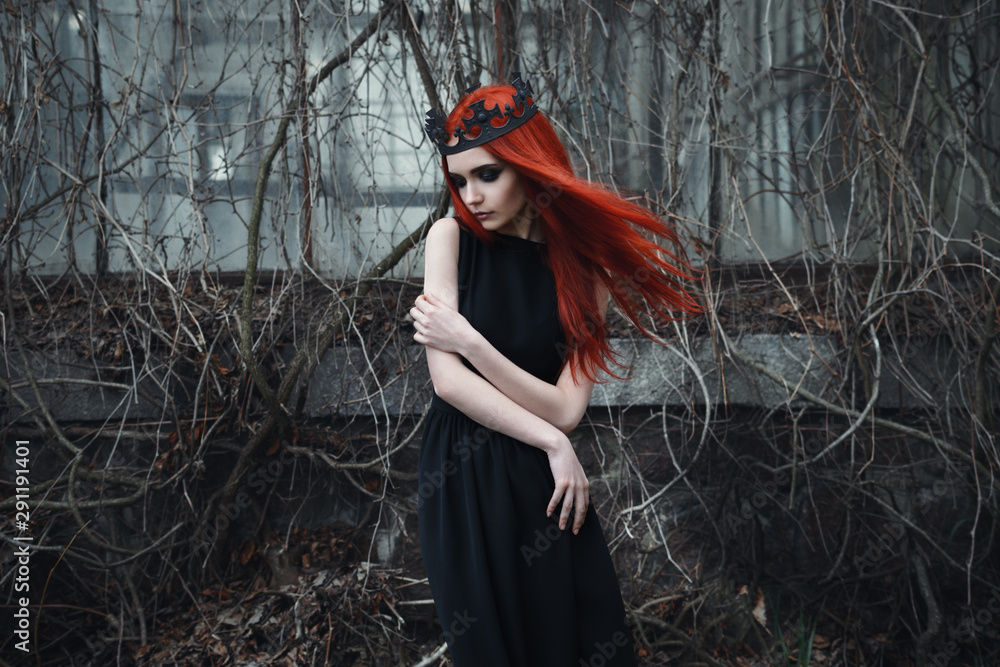 A fragile sad goth girl with red hair in a crown and a black dress ...