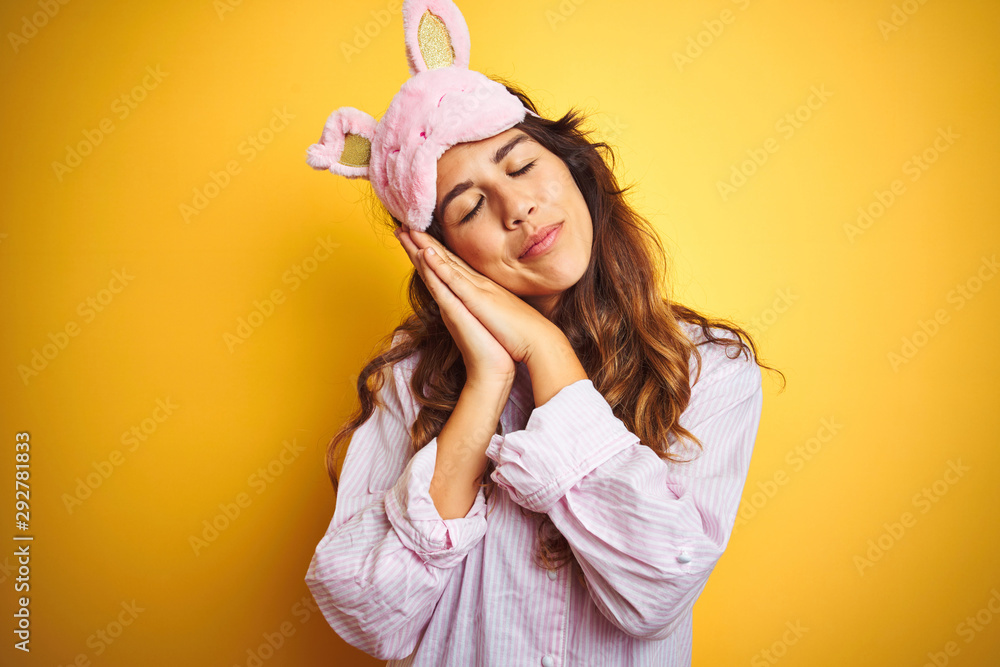 Young woman wearing pajama and sleep mask standing over yellow isolated ...