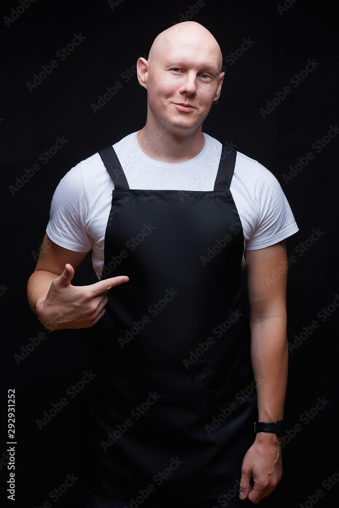 Foto de Strong handsome smiling bald guy wearing black apron pointing ...