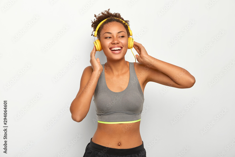 Joyful dark skinned girl enjoys loud music in headphones, has athletic ...