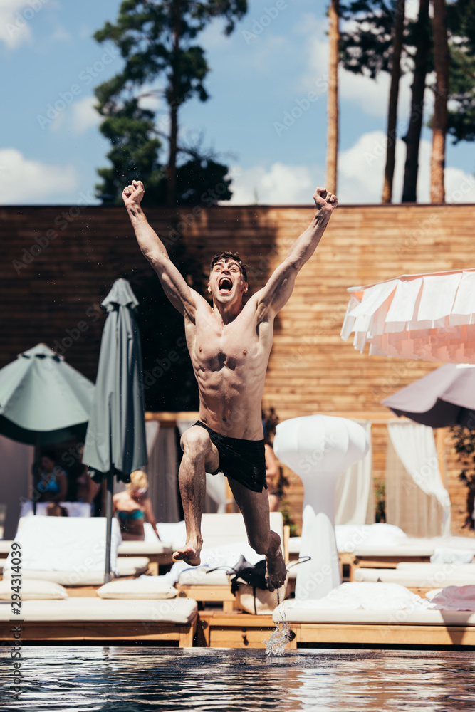 excited young man screaming while jumping in swimming pool with raised ...