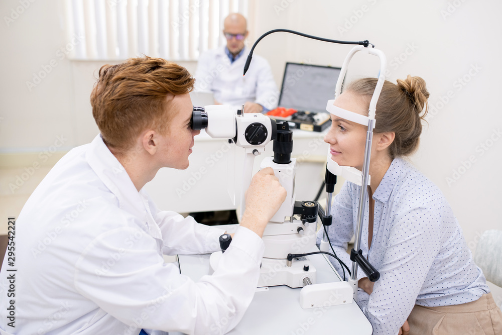 Young optometrist looking through lens of eyesight check-up equipment ...