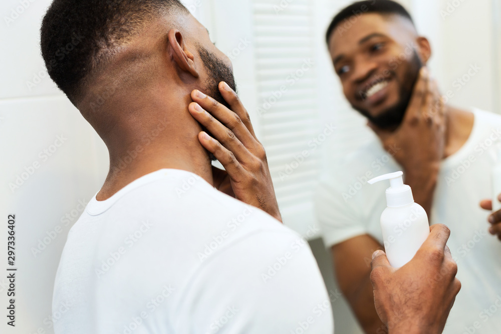 Foto de Young black man applying moisturizer on beard in bathroom do Stock | Adobe Stock