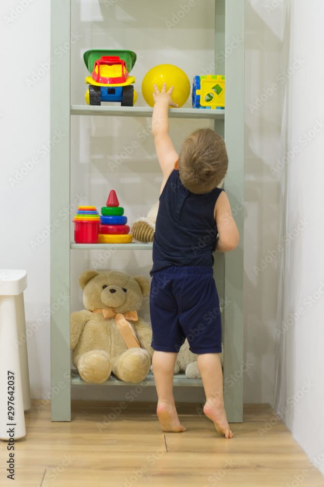 Toddler boy standing near rack and reaching for toys on shelves Stock ...